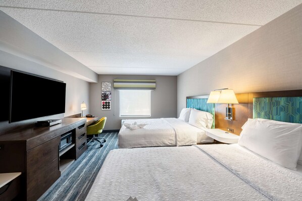 Quarto de hotel com duas camas, televisão de tela plana, escrivaninha com cadeira, janela e banheiro com espelho e porta-toalhas.