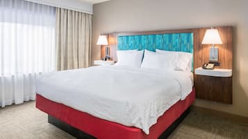 Quarto de hotel com uma cama grande, duas mesas de cabeceira com luminárias, uma cabeceira de madeira e vista para o exterior através de cortinas finas.