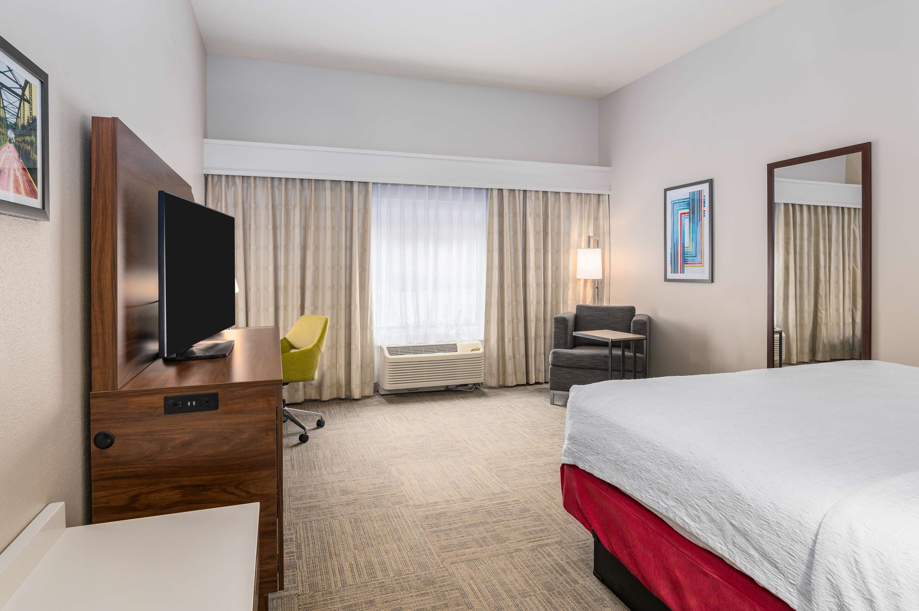 Quarto de hotel com cama, secretária com televisão, cadeira, abajur e uma grande janela com cortinas.