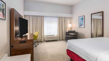 Quarto de hotel com cama, secretária com televisão, cadeira, abajur e uma grande janela com cortinas.