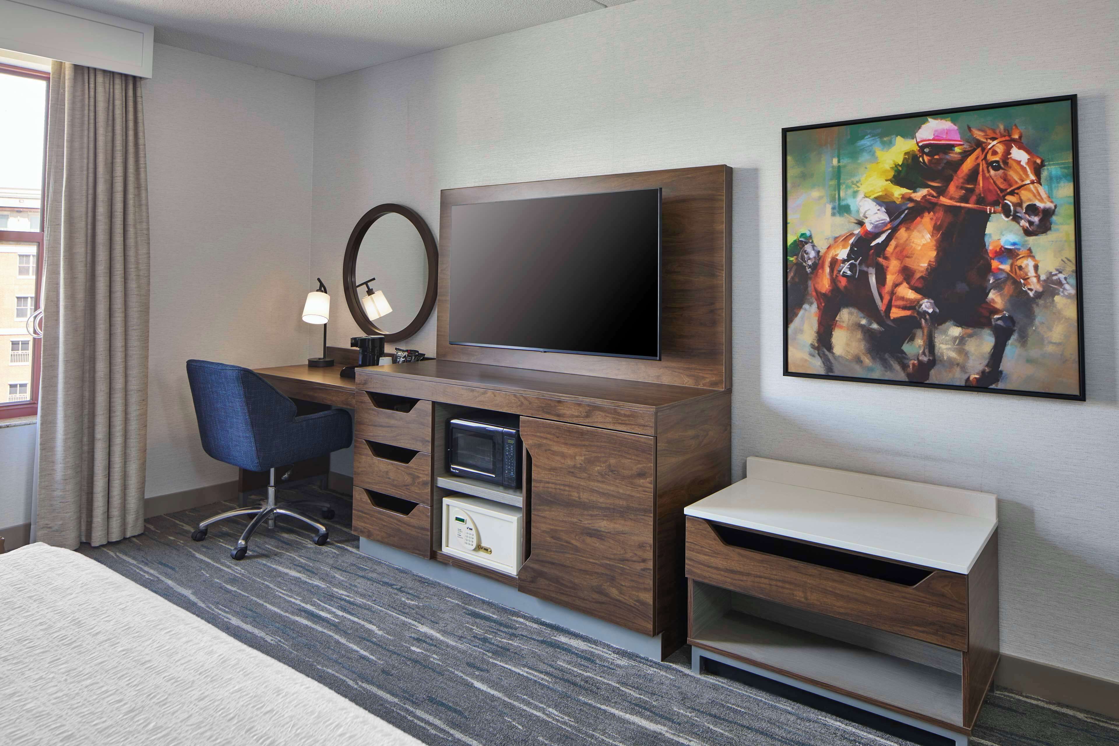 Quarto de hotel com uma televisão de tela plana grande, uma escrivaninha com cadeira, micro-ondas, um cofre pequeno, mesa de cabeceira e uma pintura de cavalos na parede.