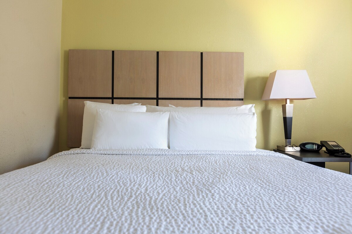 Uma cama bem arrumada com colcha branca texturizada, cabeceira de madeira com painéis horizontais e mesa de cabeceira com abajur e telefone.