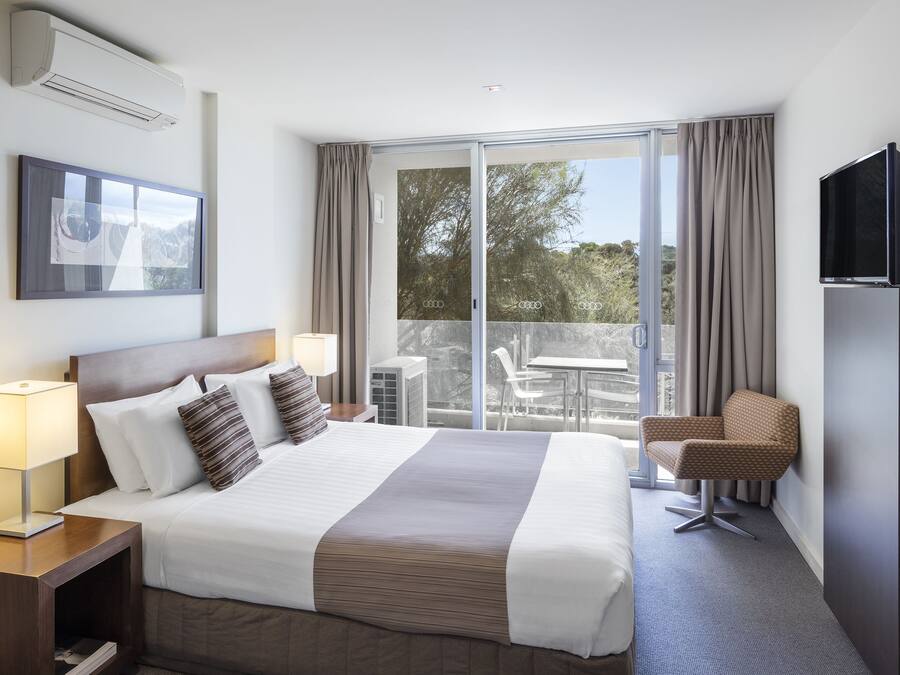 Quarto de hotel com uma cama grande, televisão, cadeira e varanda com mesa e cadeiras.
