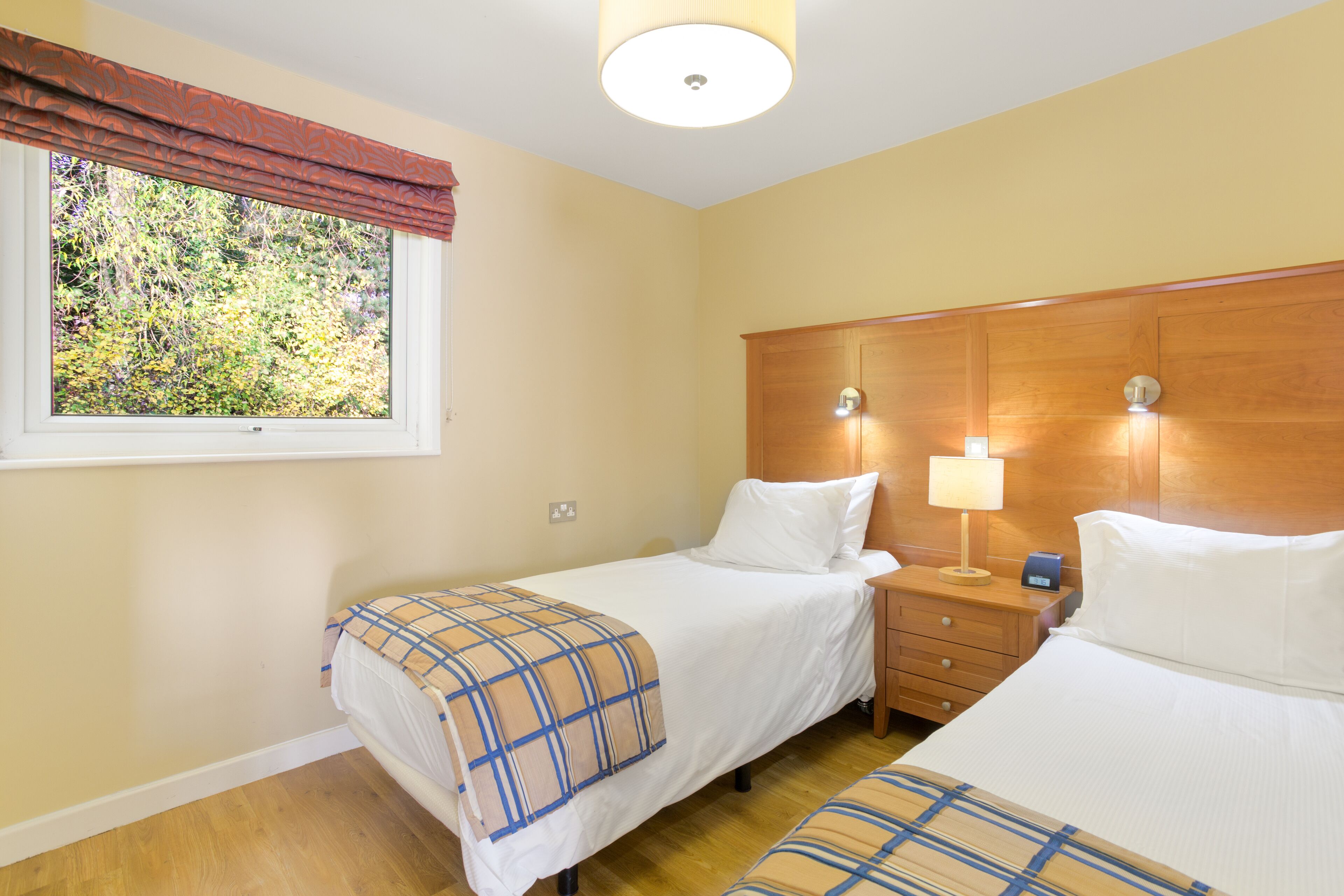 Quarto de hotel com duas camas de solteiro, cabeceira de madeira, luminárias de cabeceira, janela com vista para árvores e piso de madeira.