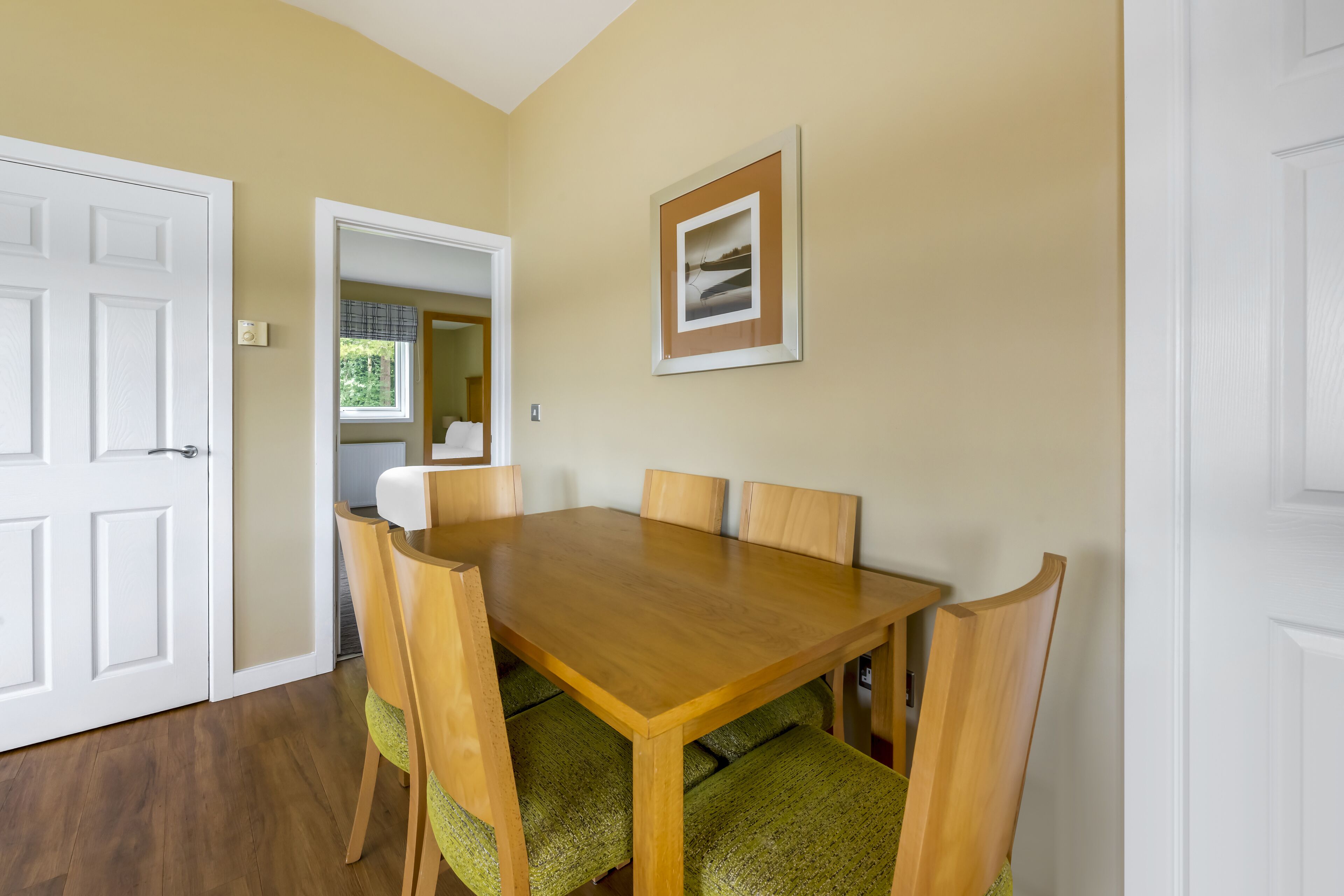 Uma área de refeições com mesa e cadeiras de madeira, um quadro na parede e uma porta à esquerda.