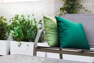 Uma cadeira de metal branca com dois apoios, um verde e um azul-turquesa, em ambiente externo com plantas ao fundo.