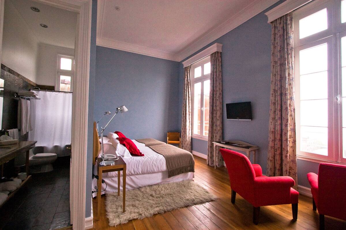 Quarto com cama, escrivaninha, cadeira, televisão e banheiro visível por uma porta aberta.