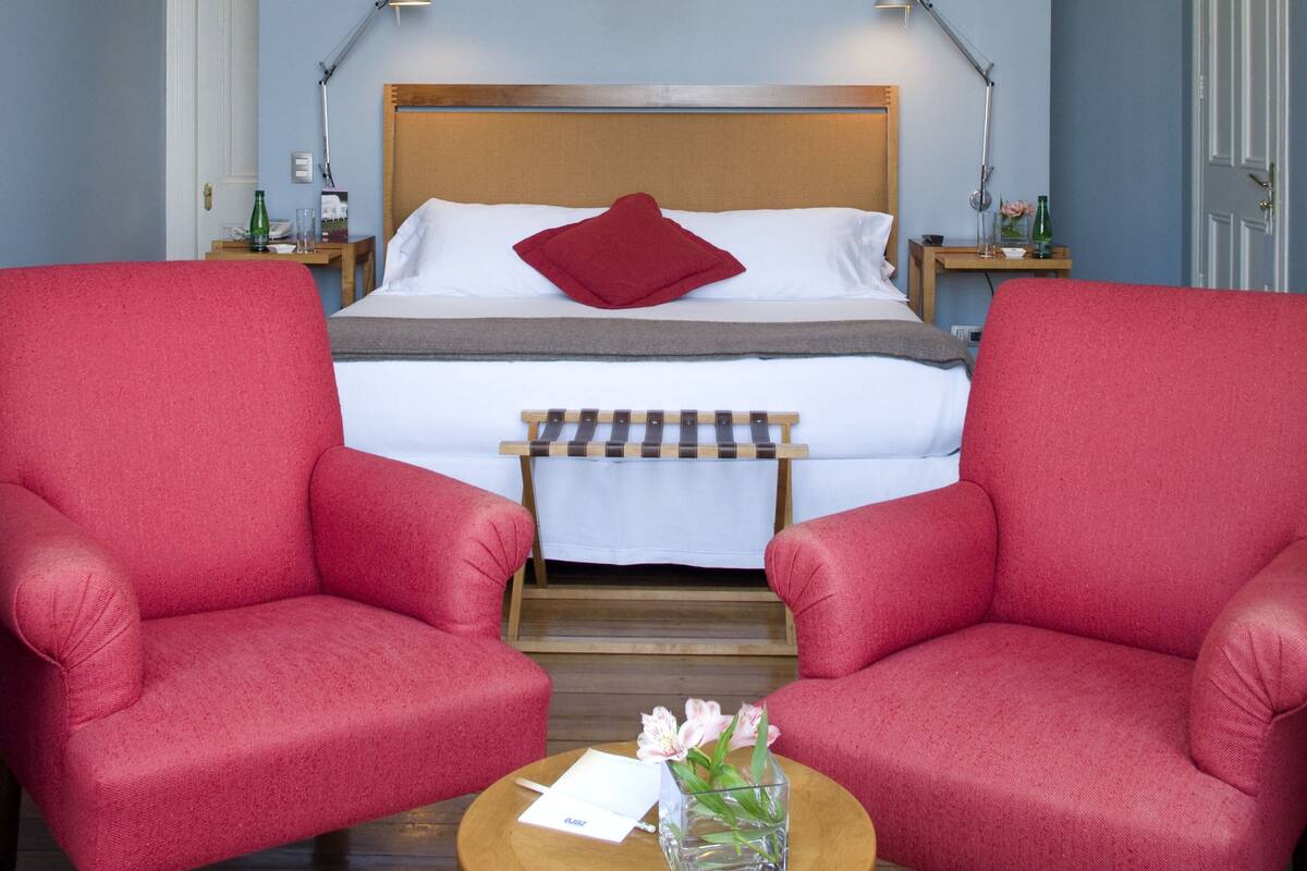 Quarto de hotel com uma cama grande, duas poltronas vermelhas, uma mesa de centro de madeira e um lustre.