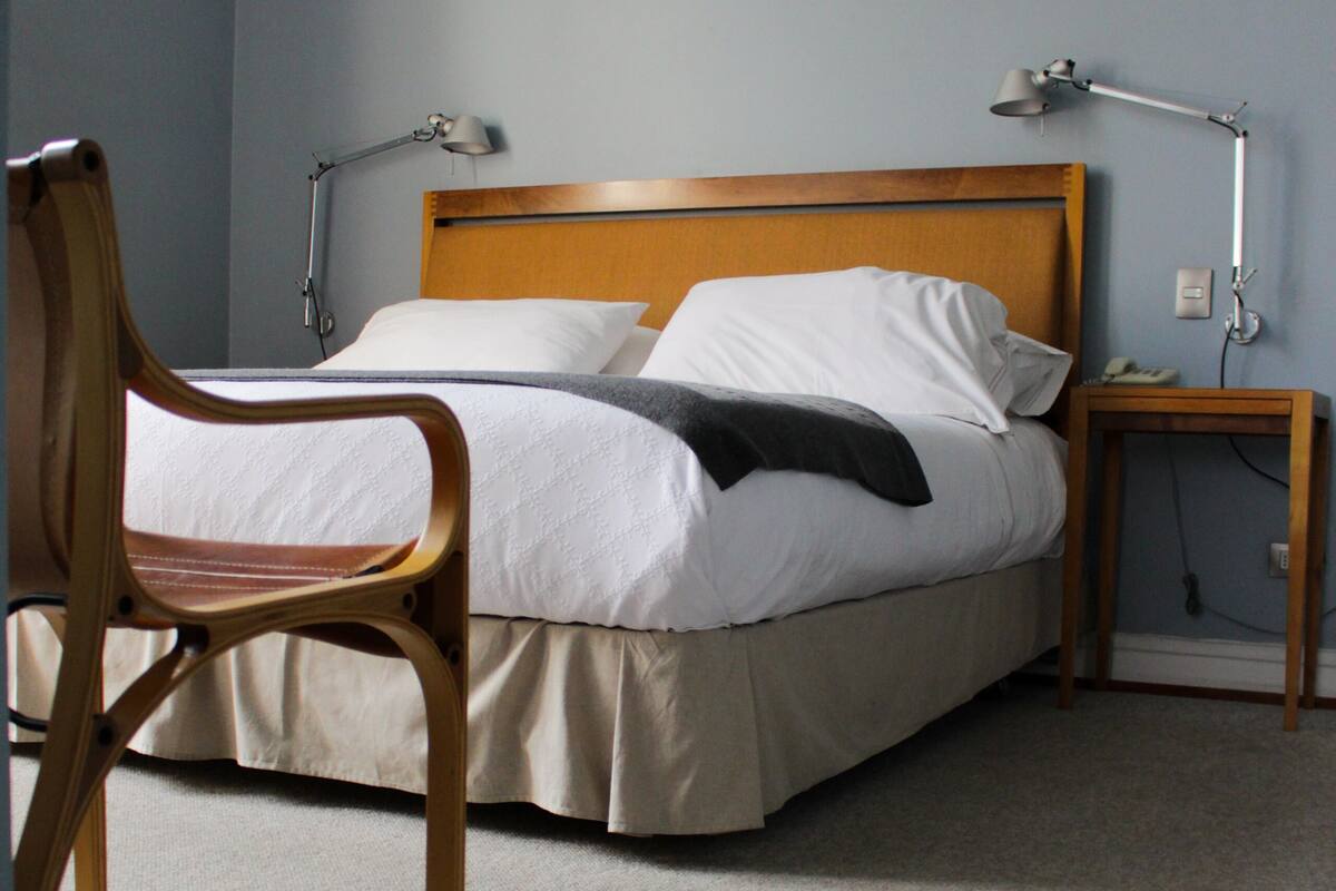 Uma cama bem arrumada com cabeceira de madeira, duas luminárias de apoio e uma cadeira em um quarto com paredes azul claro e piso coberto de carpete.