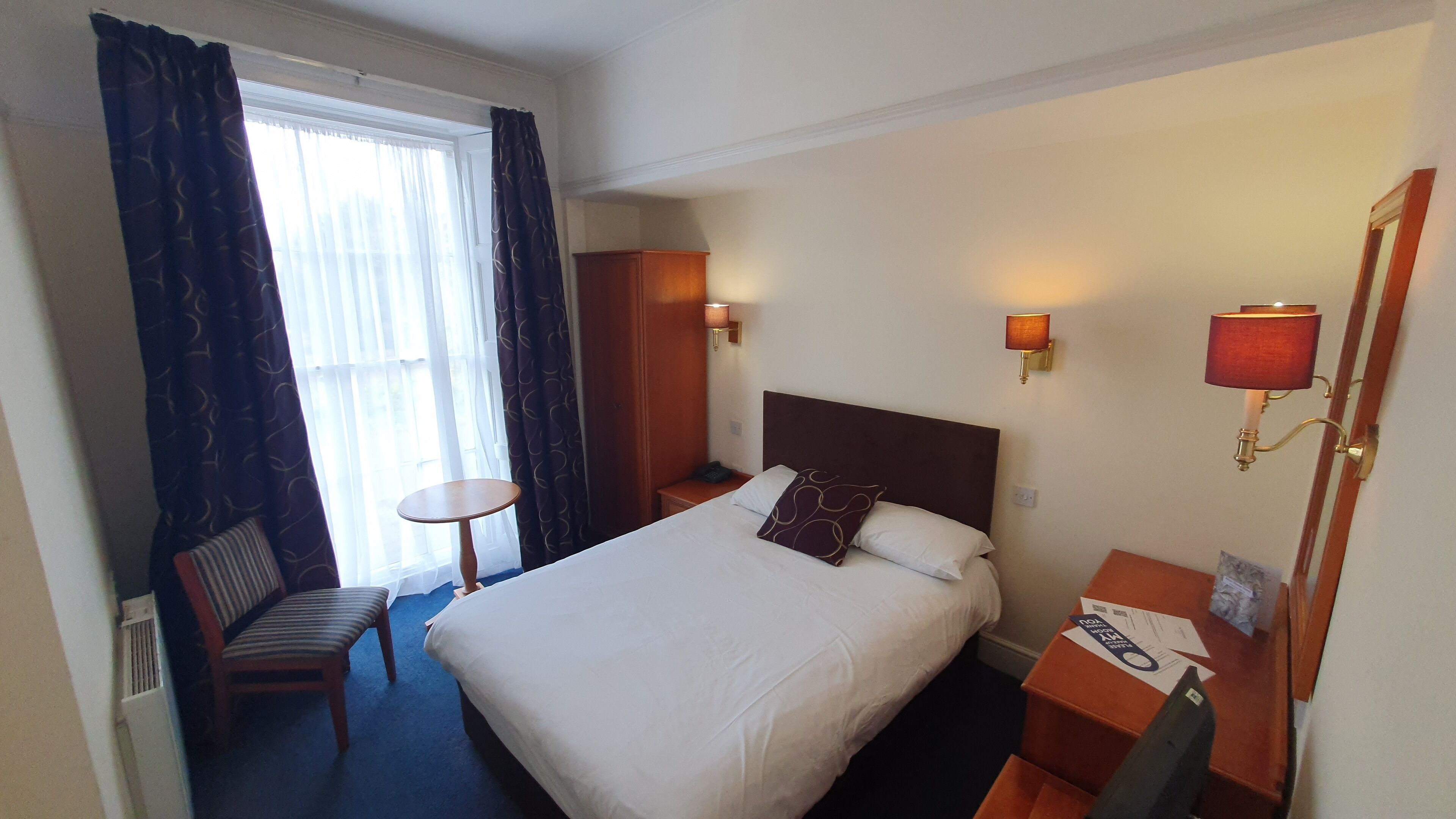 Quarto de hotel com uma cama grande, uma cadeira, uma mesa pequena e duas luminárias fixadas na parede.