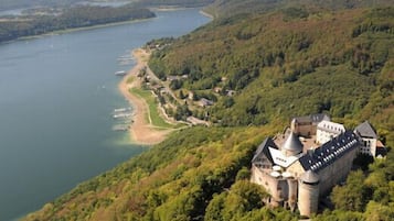 Um castelo com fosso, rodeado por uma floresta densa e um lago.