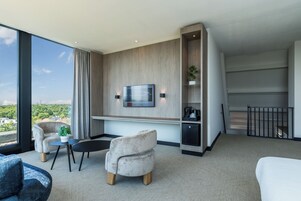 Quarto de hotel moderno com uma janela grande, televisão de tela plana na parede, uma pequena mesa de jantar, duas poltronas, um sofá e uma cozinha planejada com cafeteira.