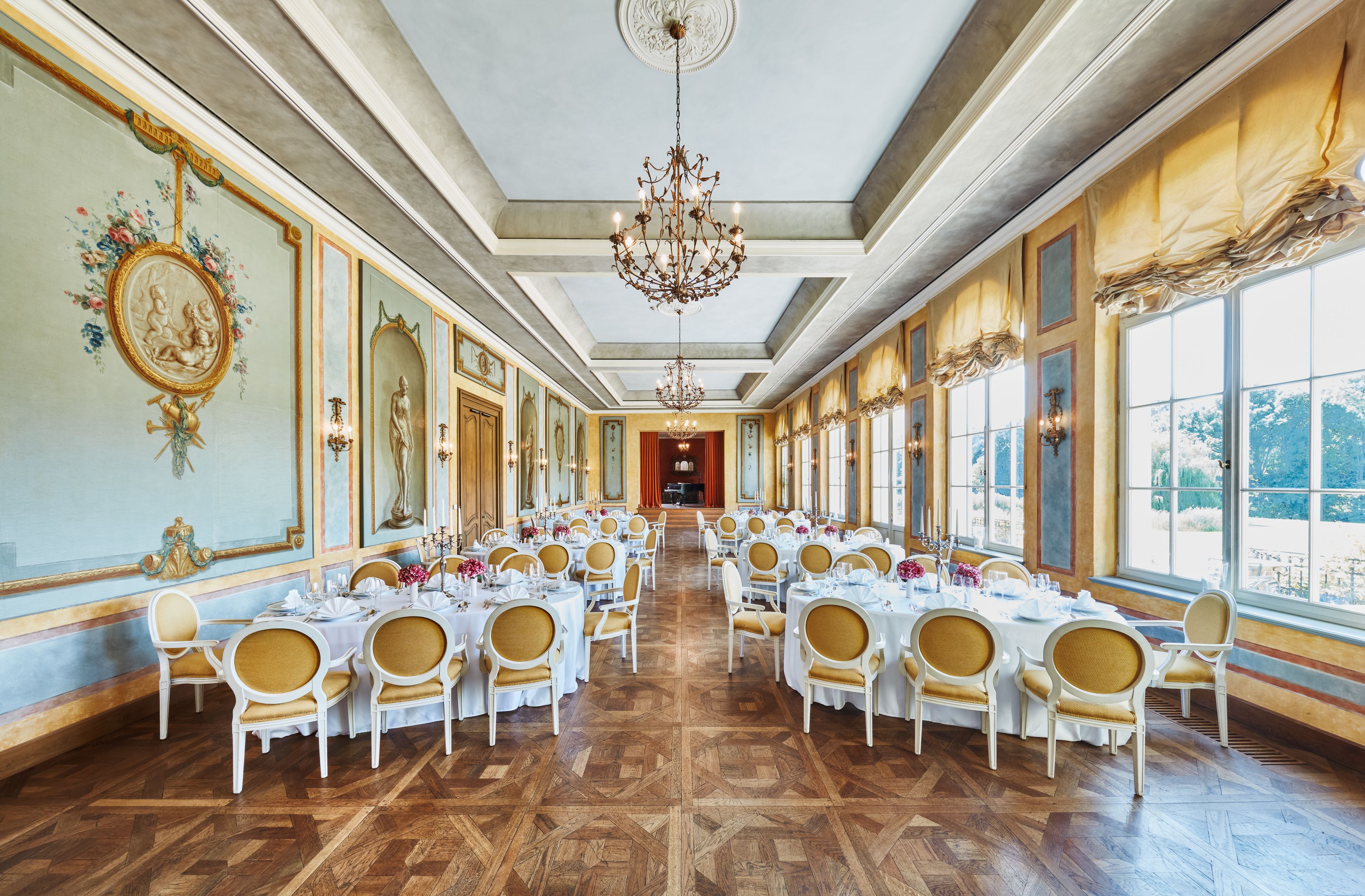 Salão de banquetes com longas mesas preparadas para refeições, lustres e grandes janelas.