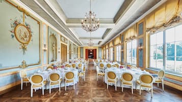 Salão de banquetes com longas mesas preparadas para refeições, lustres e grandes janelas.