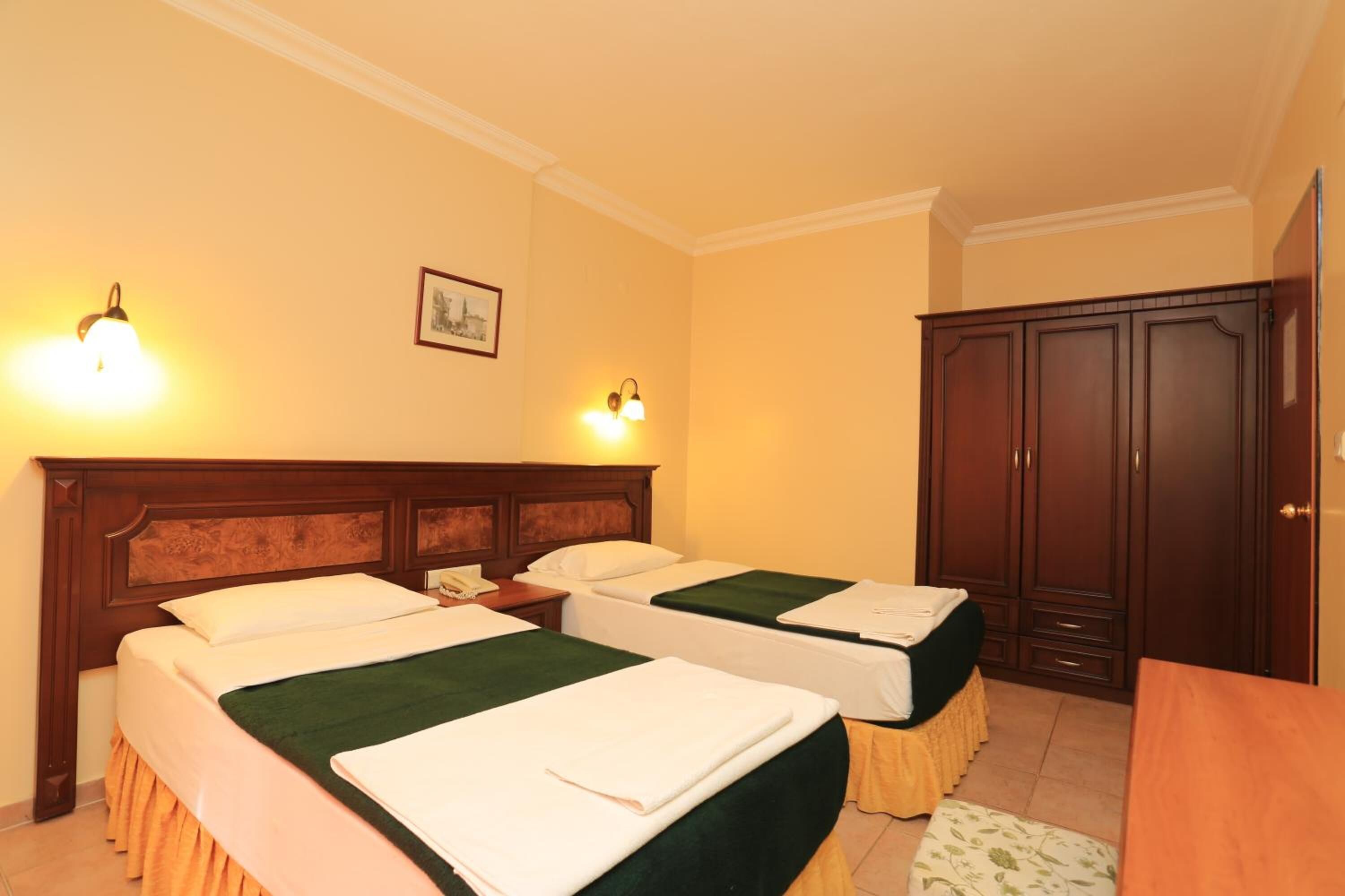 Quarto de hotel com duas camas, um guarda-roupa de madeira e duas luminárias fixadas na parede.