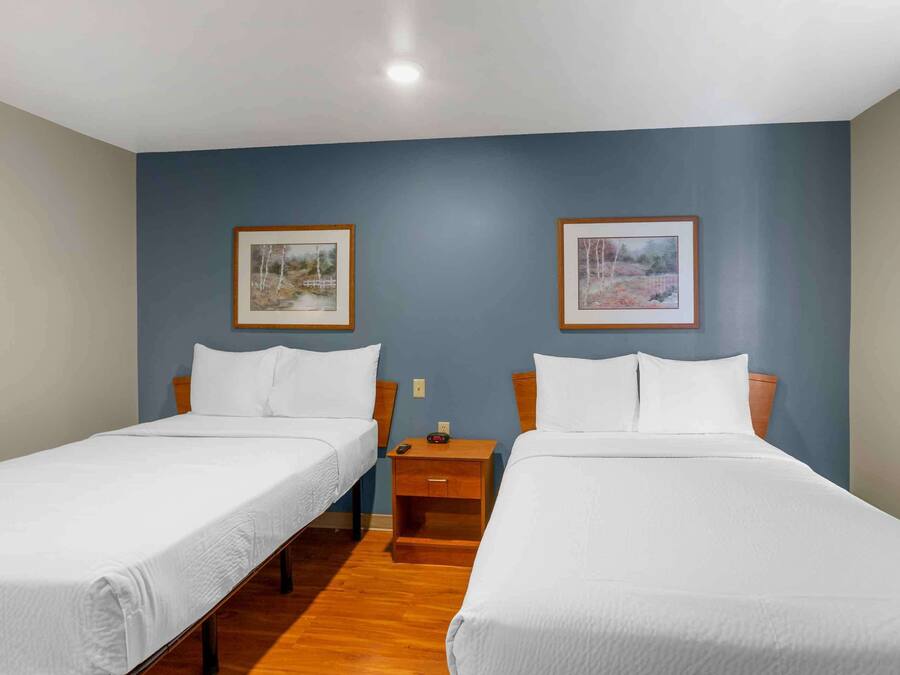 Quarto de hotel com duas camas de solteiro, uma mesinha de cabeceira de madeira e duas molduras na parede.