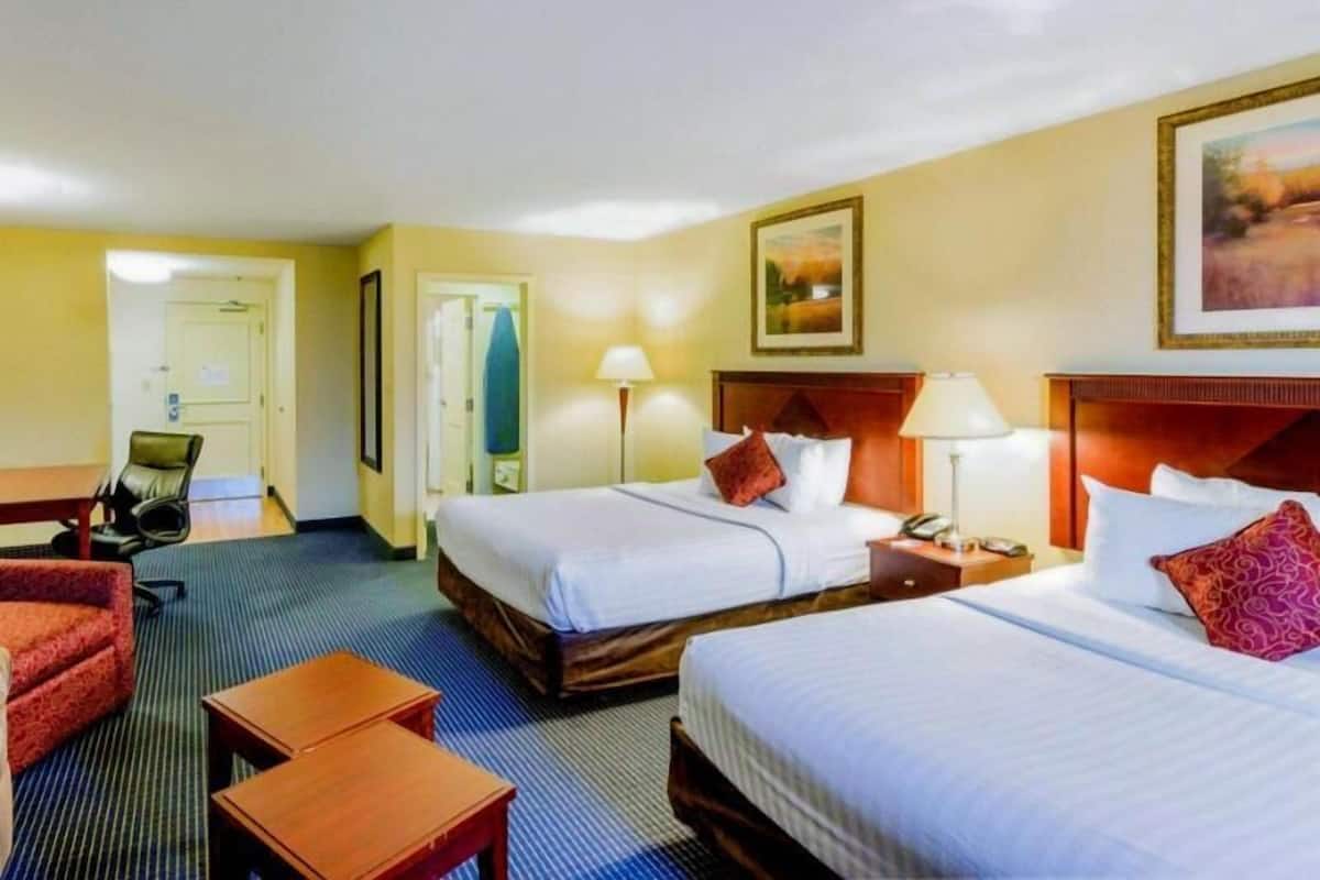 Quarto de hotel com duas camas, uma escrivaninha, uma cadeira e um abajur.