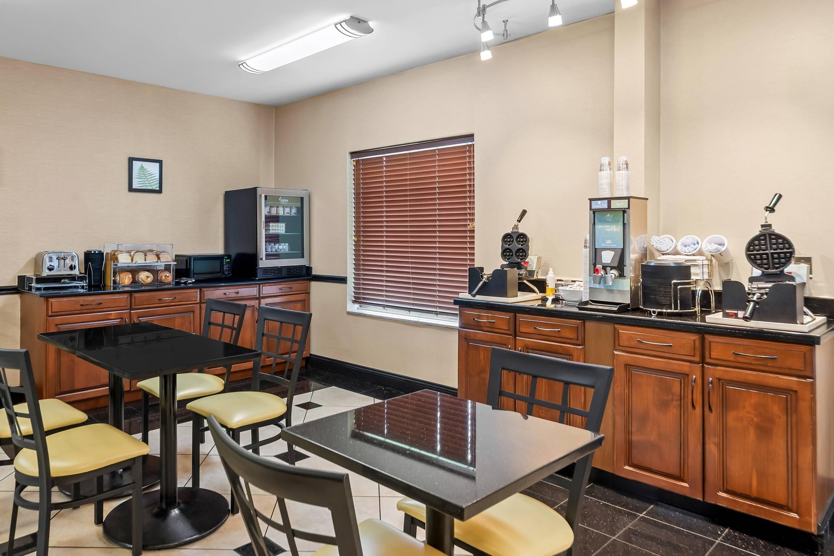 Quarto de hotel com área de jantar, cozinha compacta e janela com persianas.