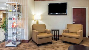 Quarto de hotel com duas poltronas bege, uma pequena mesa de apoio, uma vitrine de vidro com recordes esportivos, uma TV de tela plana montada na parede e uma porta com moldura de madeira.