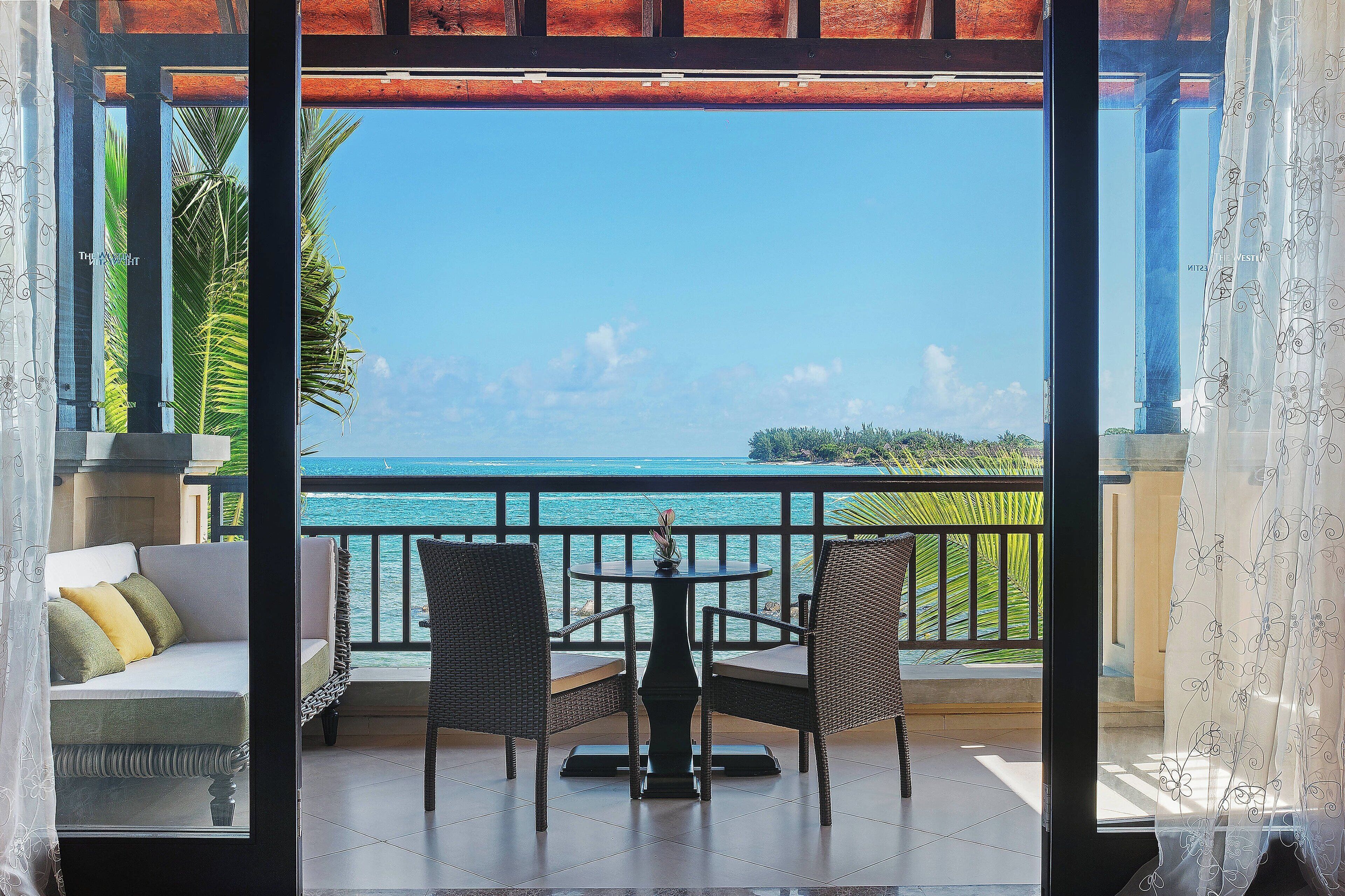 Uma varanda com vista para o mar, um sofá, duas cadeiras de vime e uma pequena mesa.
