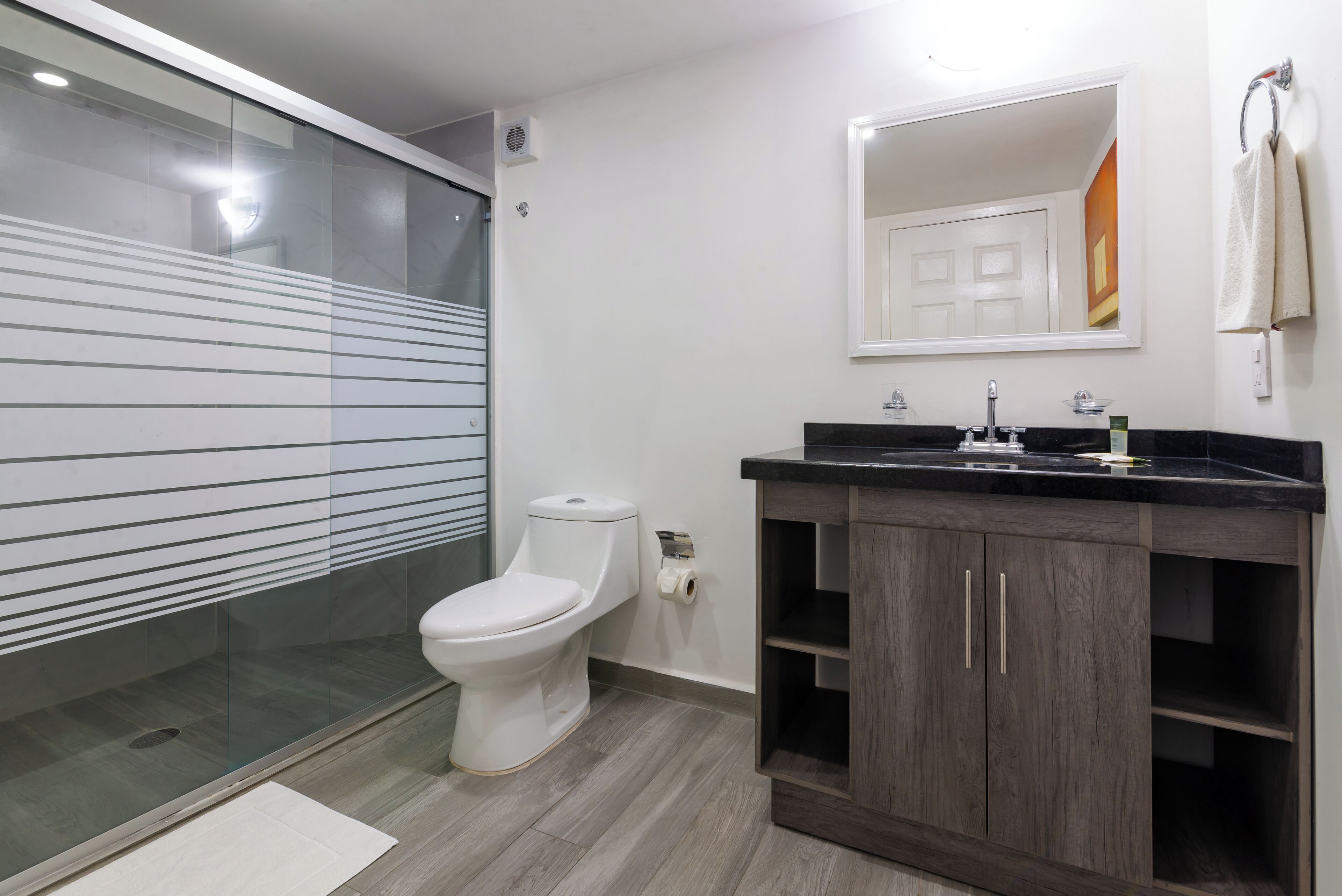 Um banheiro moderno com box de vidro, vaso sanitário branco, toucador de madeira escura com espelho e pia branca.