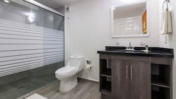 Um banheiro moderno com box de vidro, vaso sanitário branco, toucador de madeira escura com espelho e pia branca.