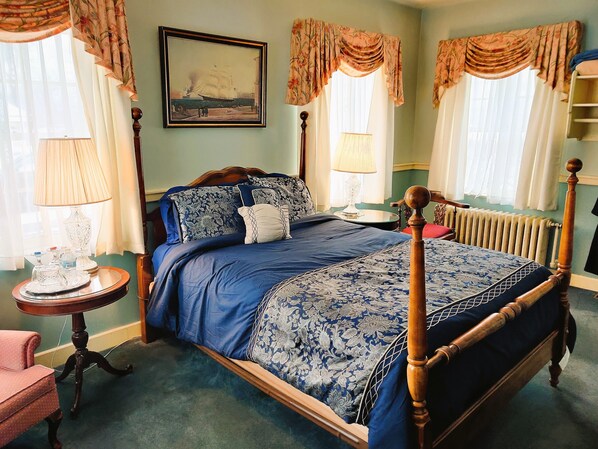 Uma cama de dossel clássica com colcha floral, uma mesa de cabeceira redonda de madeira com uma luminária e uma pintura emoldurada com tema marítimo na parede.
