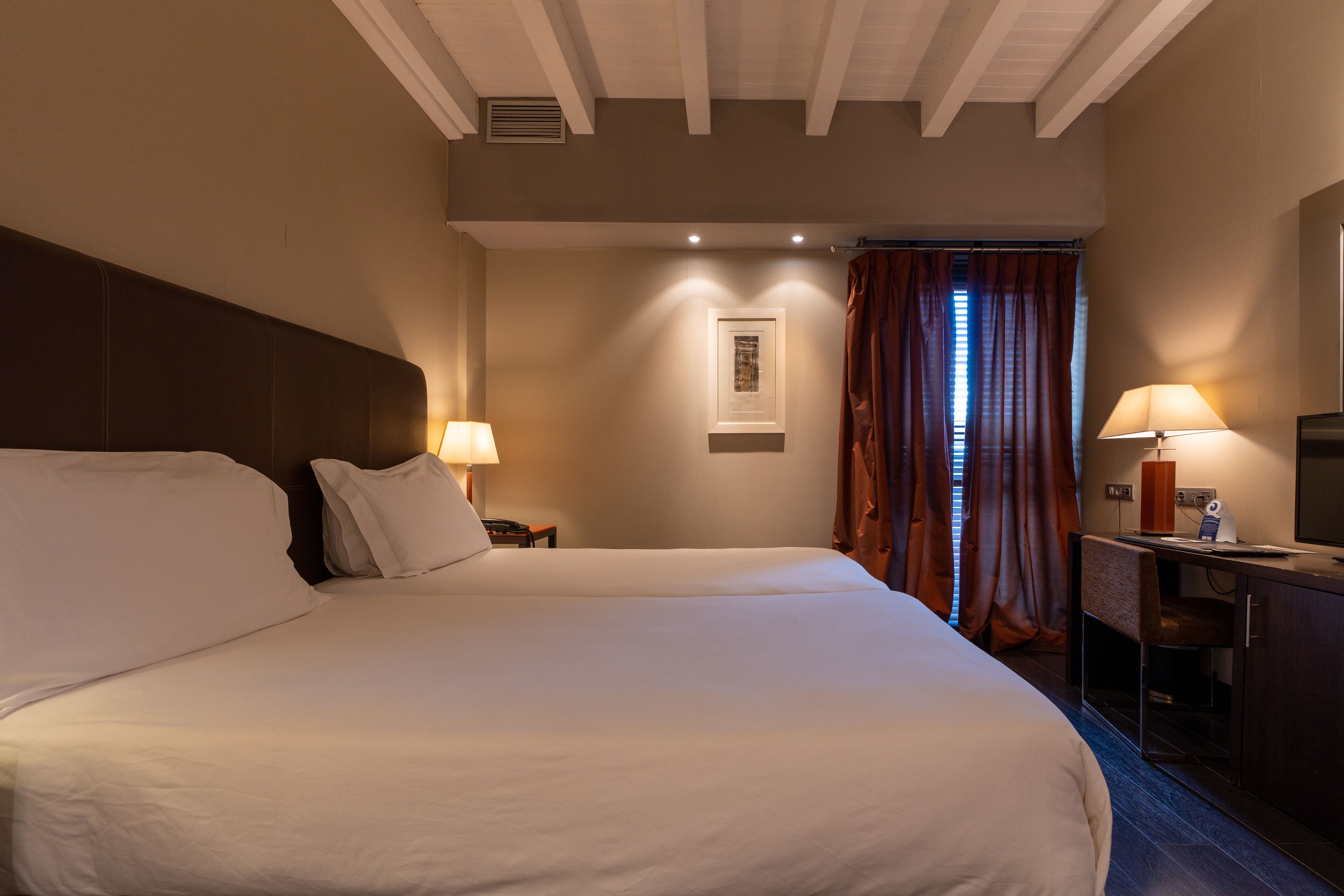 Quarto de hotel com uma cama grande, duas luminárias de cabeceira, uma escrivaninha com um computador e uma janela com cortinas.