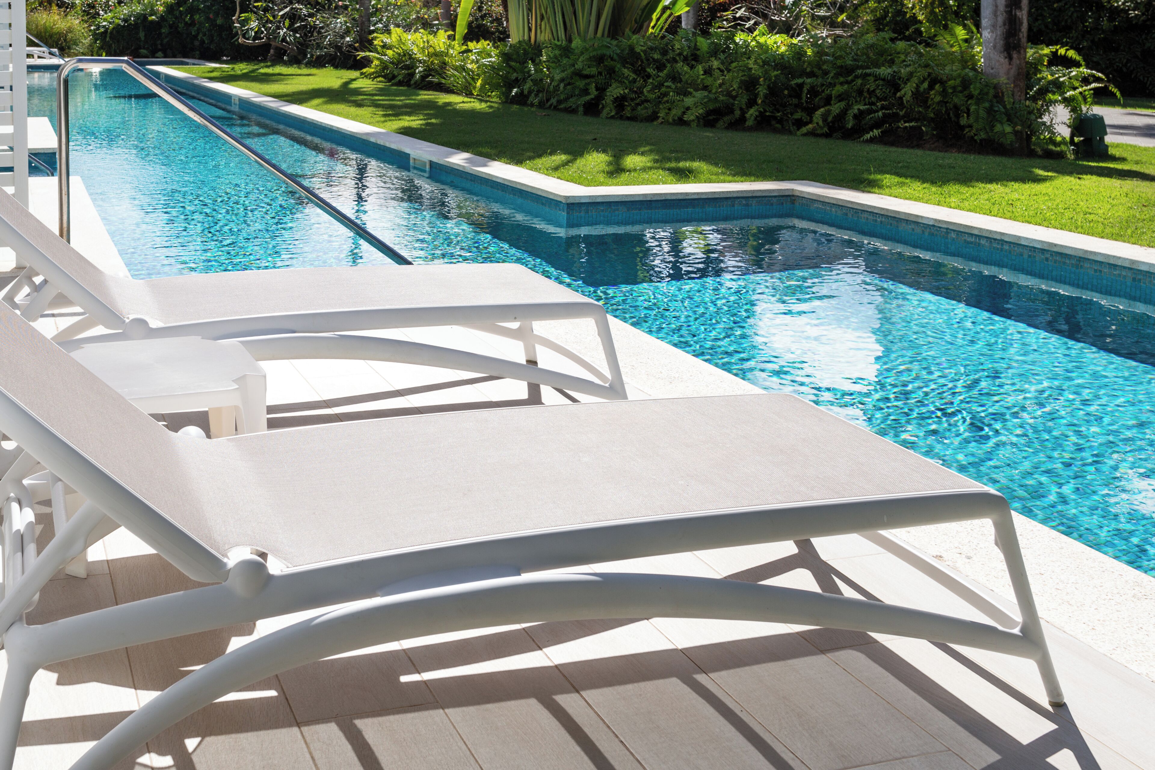Piscina com água azul cristalina, rodeada por um deck ladrilhado e cadeiras de descanso brancas.