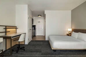 Quarto de hotel com cama, escrivaninha, cadeira e uma cozinha compacta ao fundo.