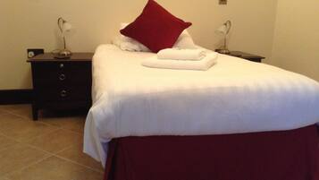 Uma cama bem arrumada com roupa de cama branca e um travesseiro vermelho, uma mesinha de cabeceira de madeira escura com uma luminária e piso ladrilhado.