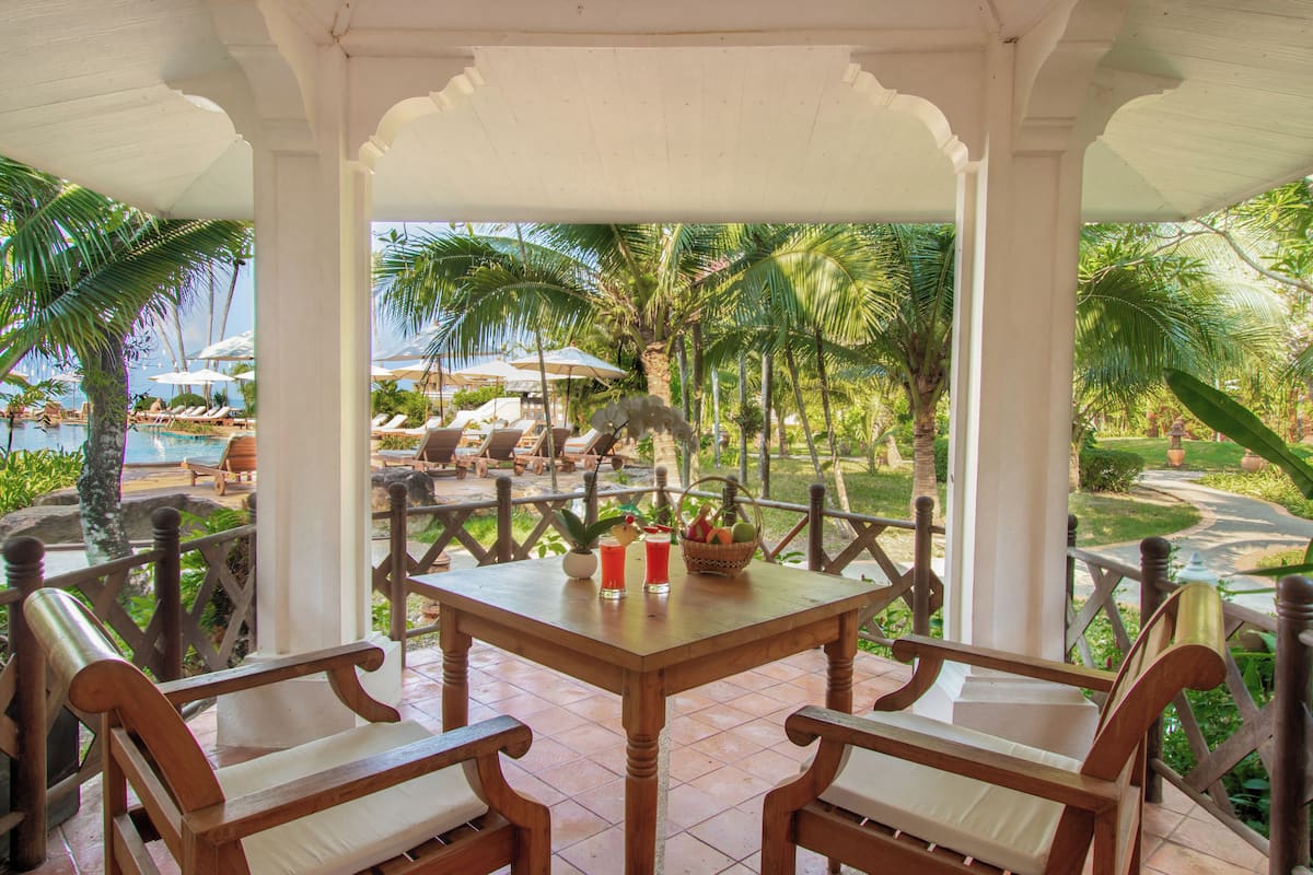 Uma área de estar coberta ao ar livre, com mesa e cadeiras, com vista para uma piscina e palmeiras.