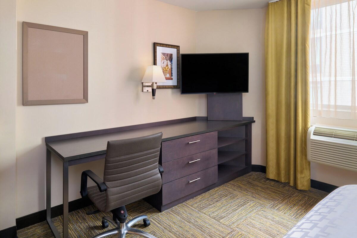 Quarto de hotel com escrivaninha, cadeira, televisão e janela com cortinas.