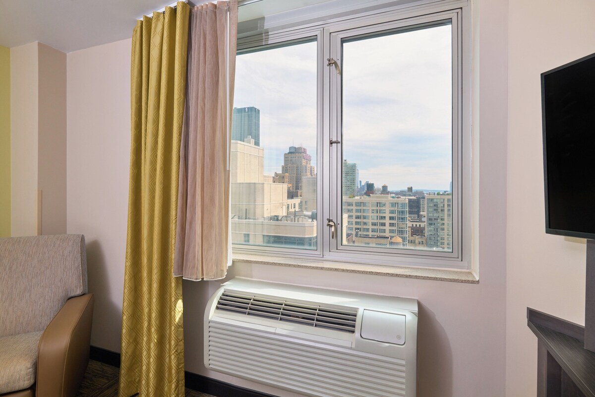 Quarto de hotel com uma janela grande, ar-condicionado fixado na parede e vista para a paisagem urbana.