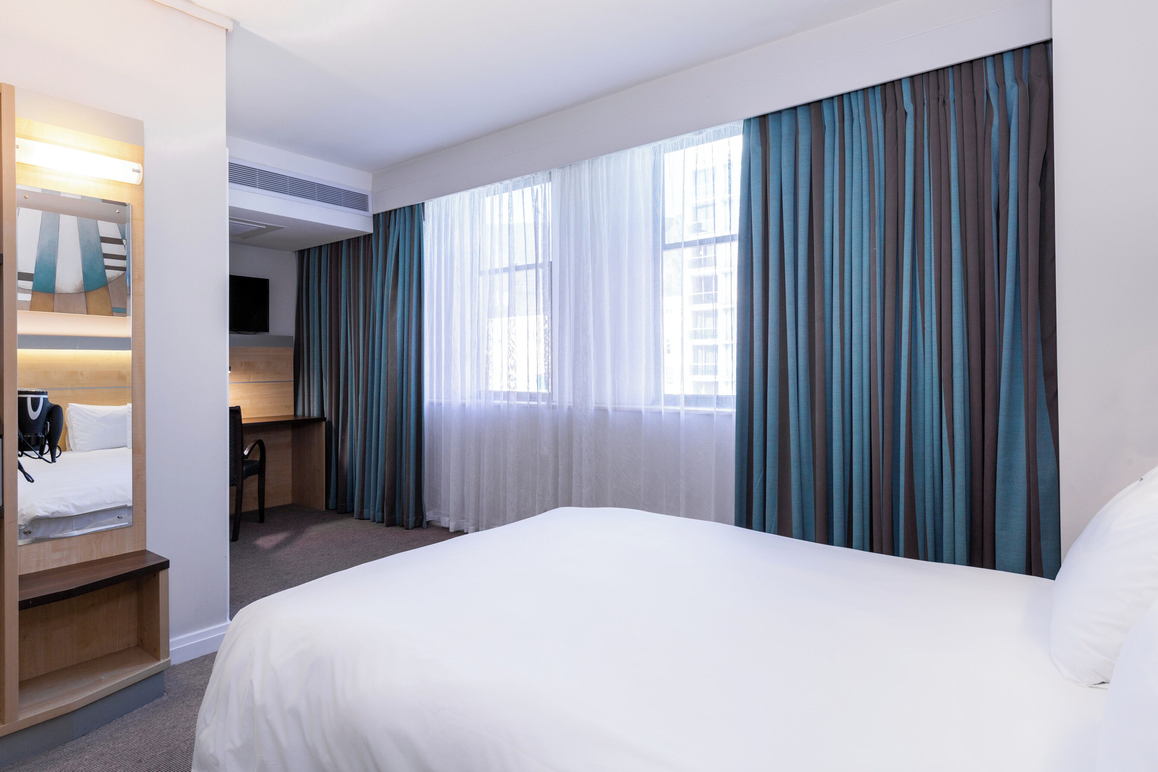 Quarto de hotel com cama, escrivaninha, cadeira, televisão e janela com cortinas.