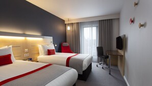 Quarto de hotel com duas camas, uma mesa, uma cadeira, televisão e janela com cortinas.