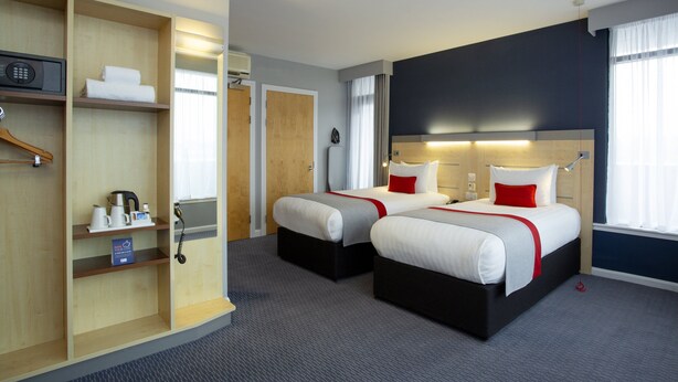 Quarto de hotel com duas camas, um guarda-roupa, uma escrivaninha e um banheiro.