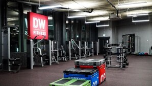 Uma academia com diversos equipamentos de exercício, um letreiro vermelho com a inscrição "DW FITNESS" e caixas PlyoSoft.