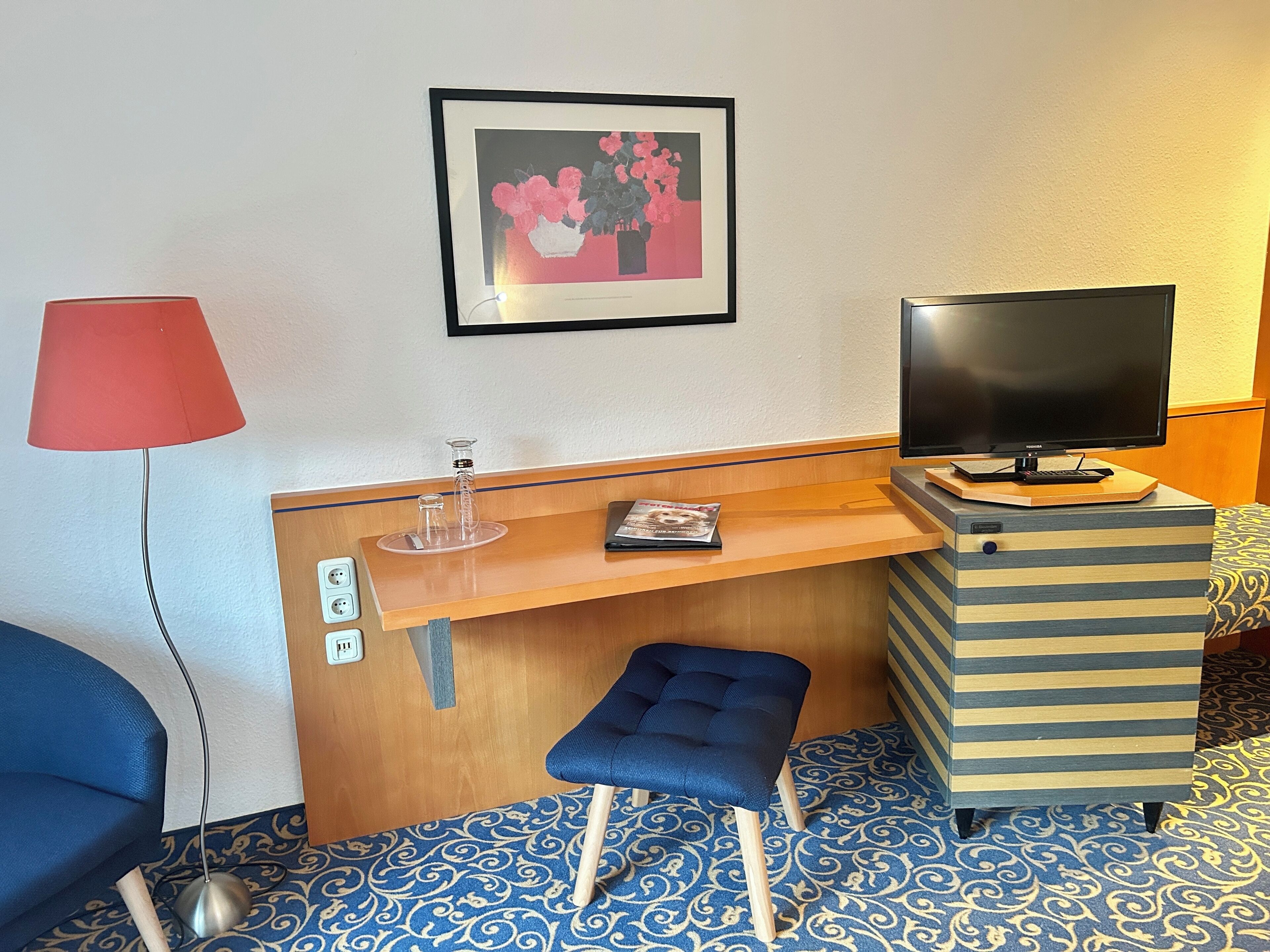 Quarto de hotel com uma escrivaninha de madeira, uma televisão sobre suporte de madeira, uma cadeira azul, um abajur vermelho e um quadro emoldurado na parede.