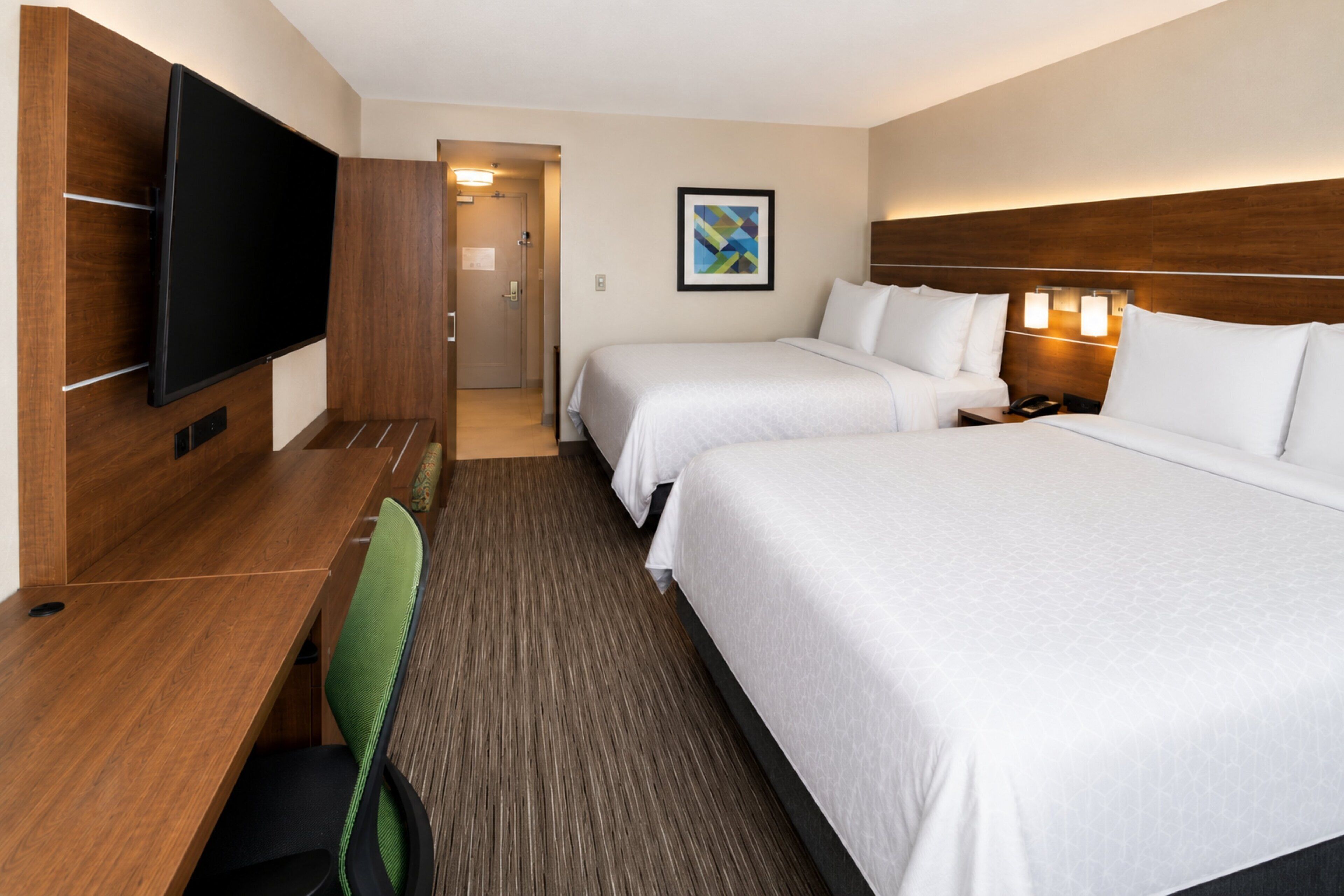 Quarto de hotel com duas camas, uma escrivaninha, uma televisão e uma porta para um corredor.