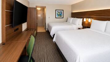 Quarto de hotel com duas camas, uma escrivaninha, uma televisão e uma porta para um corredor.