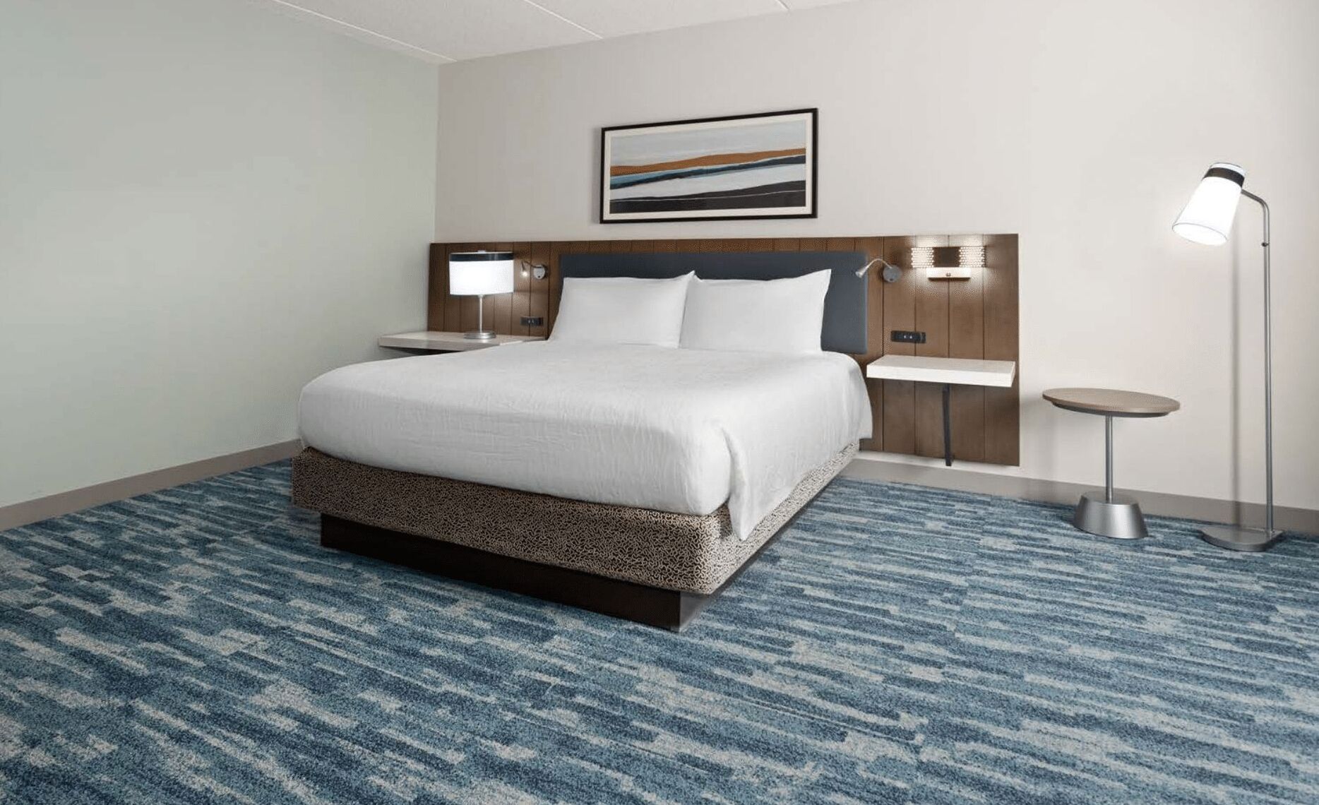 Quarto de hotel com uma cama grande, mesa de cabeceira, escrivaninha, luminária e quadro na parede.
