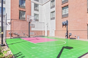 Quadra de basquete ao ar livre com superfície de jogo verde, cercada por um edifício de tijolos com várias janelas e um hidrante visível.