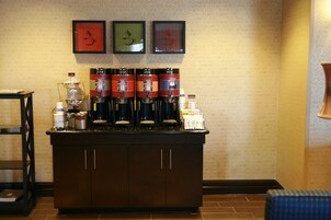 Uma estação de café com vários dispensadores, uma pequena prateleira com acompanhamentos e uma obra de arte emoldurada na parede.