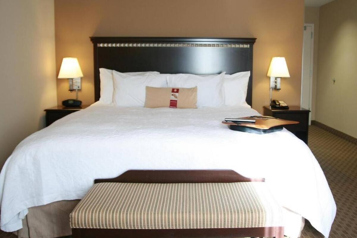 Quarto de hotel com cama grande, duas luminárias de cabeceira, mesa de cabeceira e um banco.
