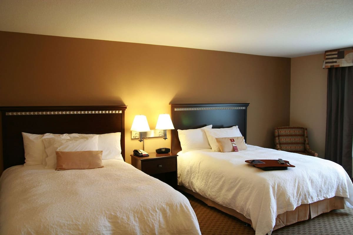Quarto de hotel com duas camas, mesas de cabeceira, abajures, uma cadeira e uma janela com cortinas.
