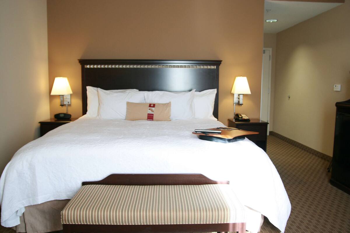 Cama bem arrumada com lençóis brancos, duas luminárias de cabeceira, uma mesinha de cabeceira de madeira e uma pequena mesa lateral com um livro.