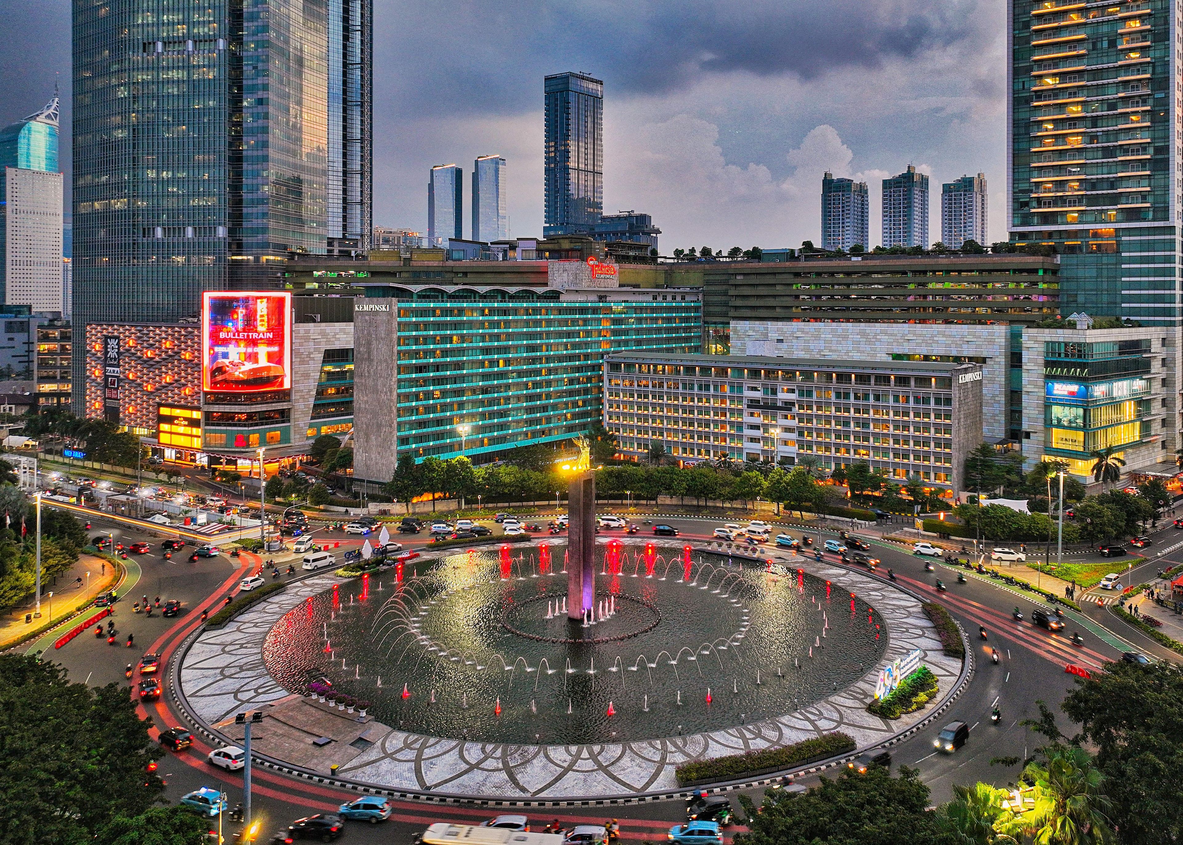 Paisagem urbana com um hotel em destaque, apresentando um letreiro grande e iluminado, cercado por edifícios altos e modernos, e uma fonte circular no centro.