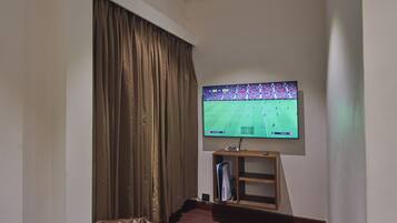 Quarto com televisão exibindo uma partida esportiva, uma poltrona de milho e uma prateleira de madeira com um console de jogos.