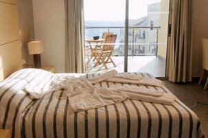 Quarto de hotel com cama, mesa e cadeiras, e vista para o mar através de uma varanda.